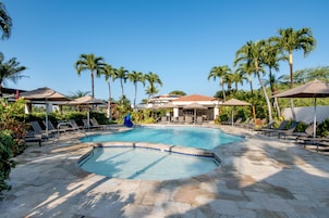 Área de piscina com espreguiçadeiras e guarda-sóis, cercada por palmeiras e um edifício com telhado de azulejos.