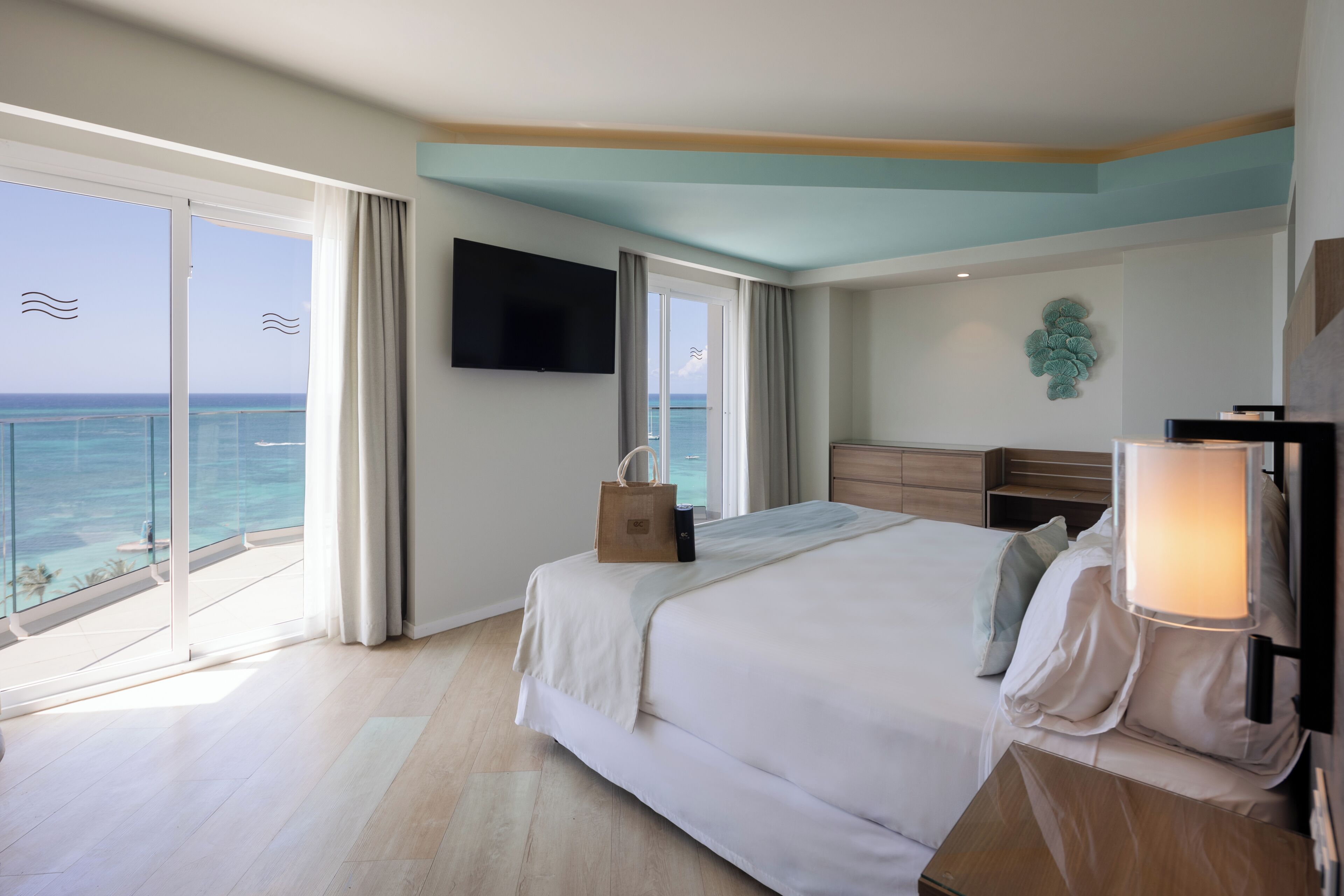 Quarto de hotel com cama, televisão, mesa de cabeceira, luminária, janela com vista para o oceano e uma obra de arte na parede.