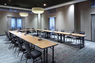 Uma sala de conferência com mesas compridas e cadeiras, grandes janelas com cortinas e um tapete estampado.