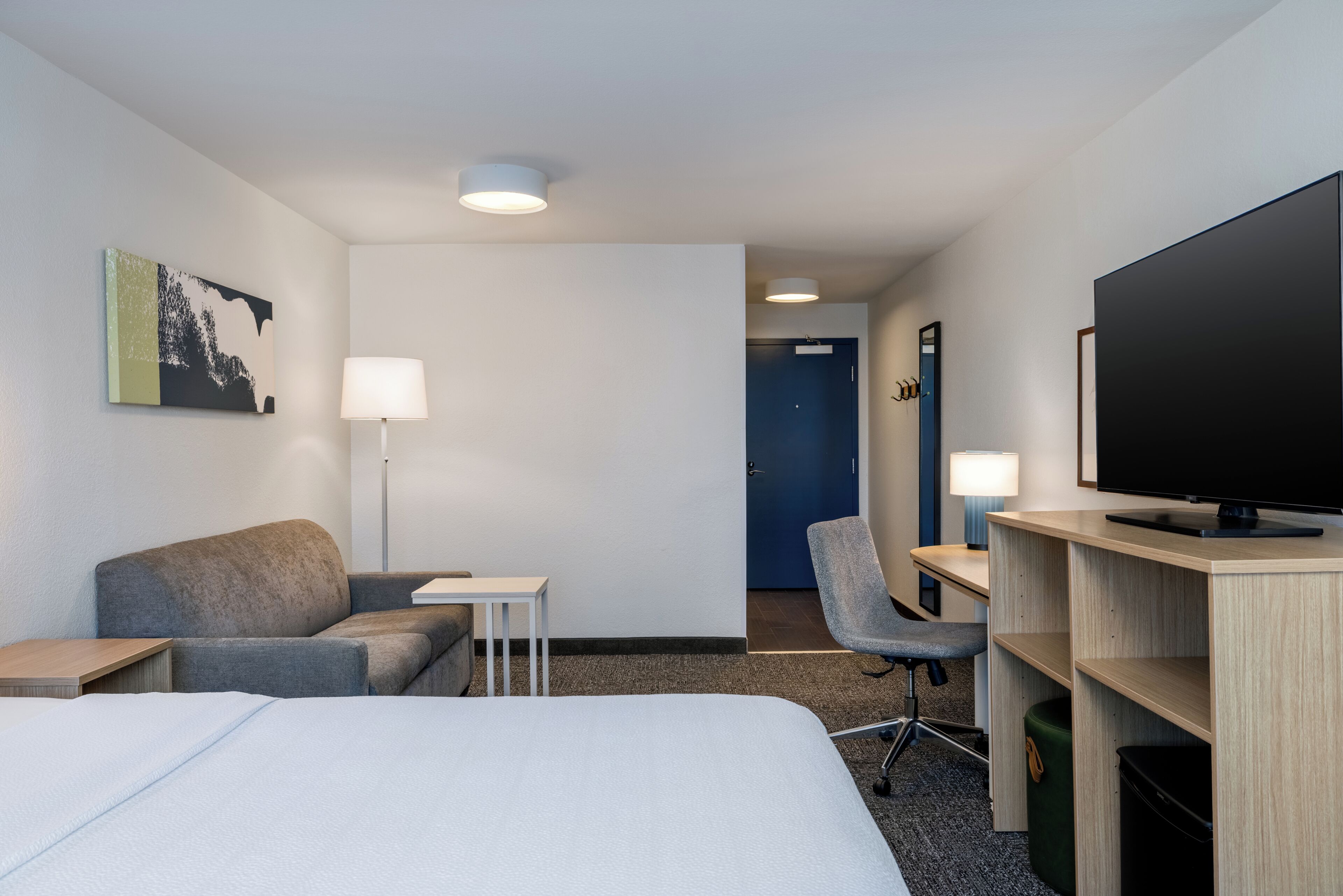 Quarto de hotel com cama, escrivaninha, cadeira, televisão e um quadro na parede.