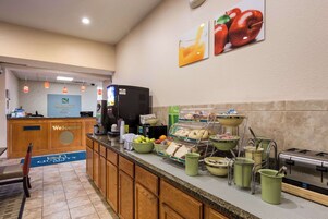 Uma área de recepção de hotel com uma placa de 'Welcome', uma máquina de café e um buffet de café da manhã com diversos alimentos.