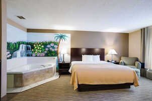 Quarto de hotel com uma cama grande, banheiro separado com banheira, pintura de uma cachoeira e palmeira, e mesa de cabeceira com abajur.