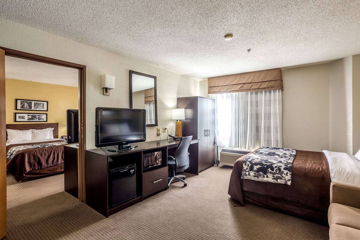 Quarto de hotel com cama, secretária com televisão, cadeira, guarda-roupa e janela com cortinas.