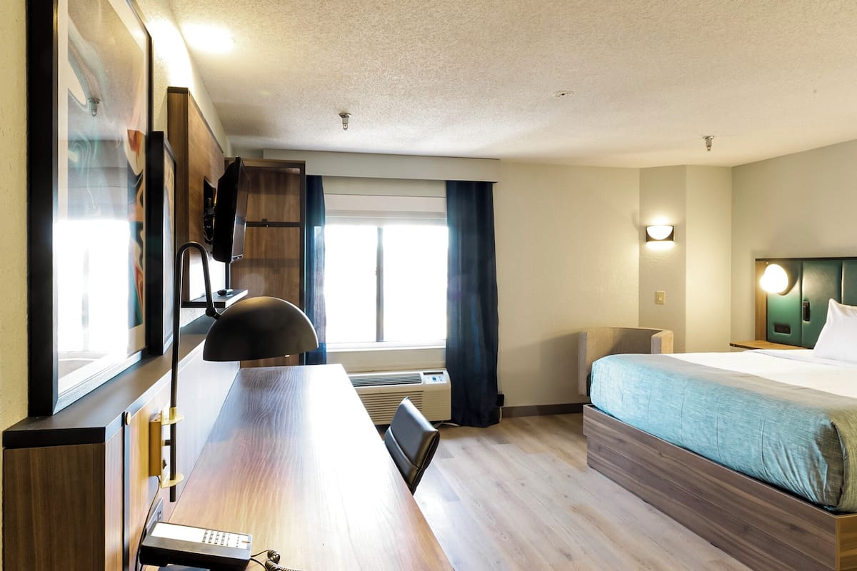 Quarto de hotel com cama, escrivaninha, cadeira e uma grande janela com cortinas.
