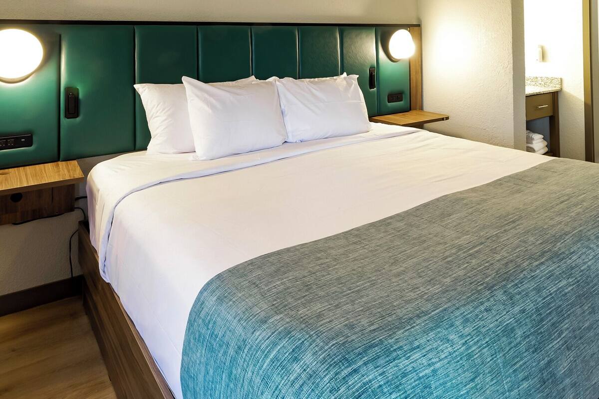 Cama bem arrumada com roupa de cama branca e manta azul-esverdeada, mesa de cabeceira de madeira com abajur, e cabeceira verde com luminárias redondas.
