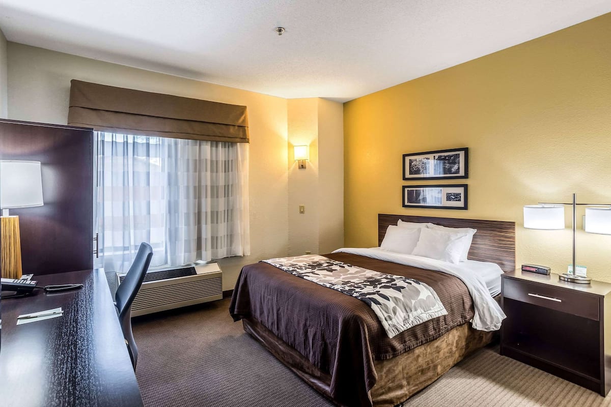 Quarto de hotel com cama, escrivaninha e cadeira, janela com cortinas e duas luminárias na parede.