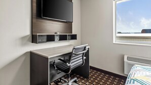 Quarto de hotel com televisão de tela plana na parede, uma escrivaninha com cadeira, uma janela com vista para edifícios e piso estampado.