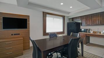 Uma sala de conferências com uma mesa grande, cadeiras, uma TV de tela plana, armários de madeira e uma área de cozinha com eletrodomésticos.