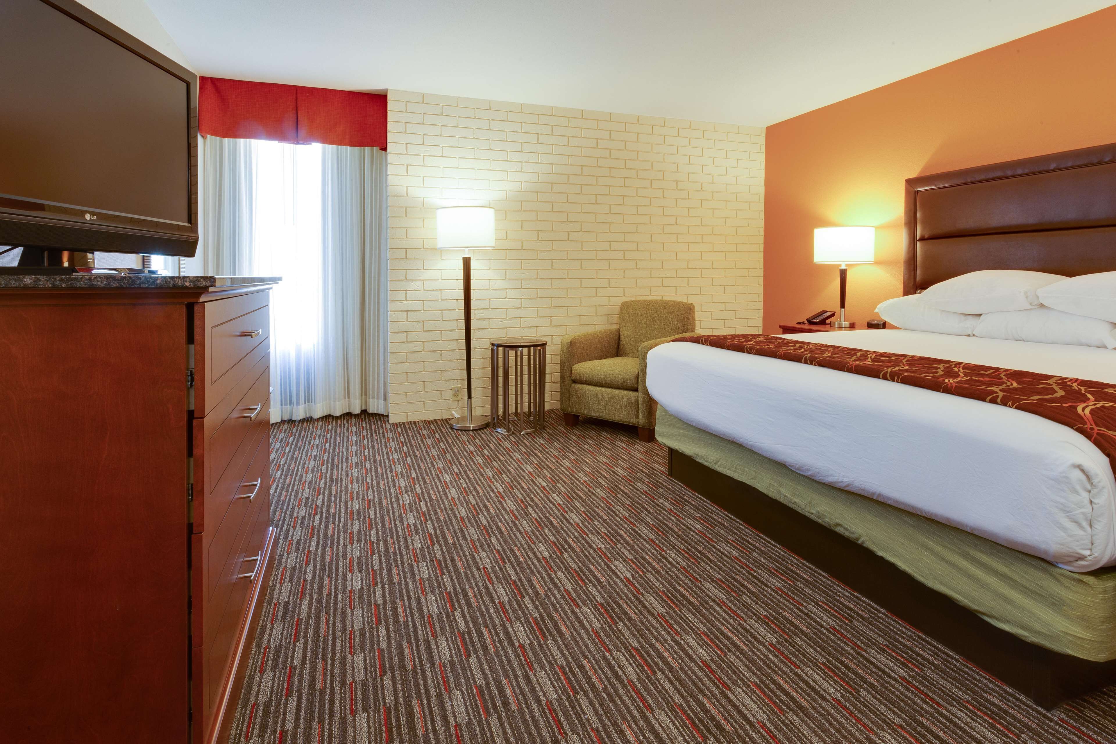 Quarto de hotel com cama, televisão, cadeira, abajur e pequena mesa.