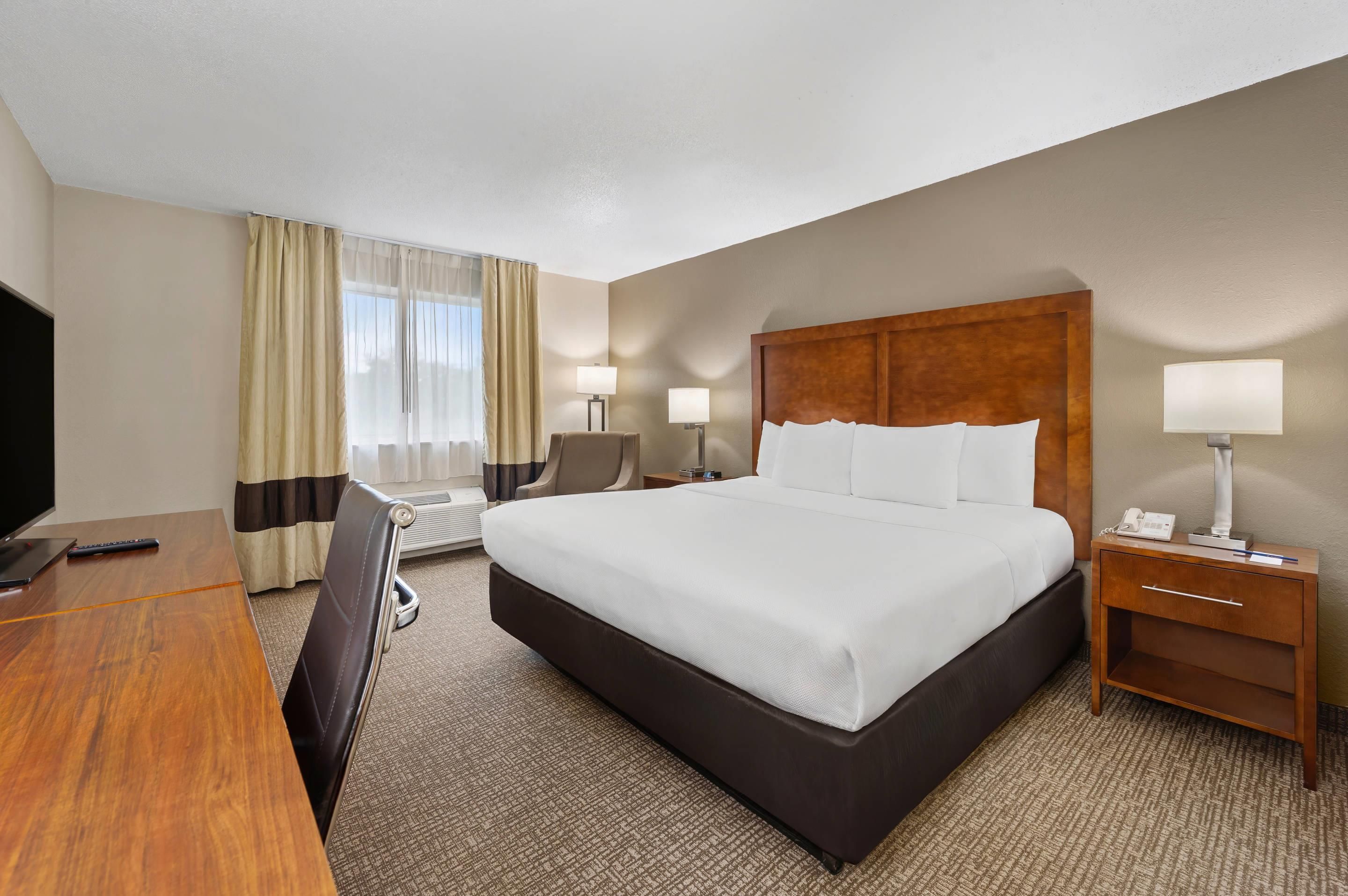 Quarto de hotel com uma cama grande, escrivaninha com cadeira, televisão e duas luminárias de cabeceira.