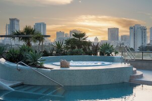 Piscina no terraço com hidromassagem, rodeada por palmeiras e um horizonte urbano ao fundo.