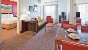 Quarto de hotel com uma cama, um sofá, uma mesa de jantar posta com pratos e talheres, uma televisão e uma cadeira vermelha.