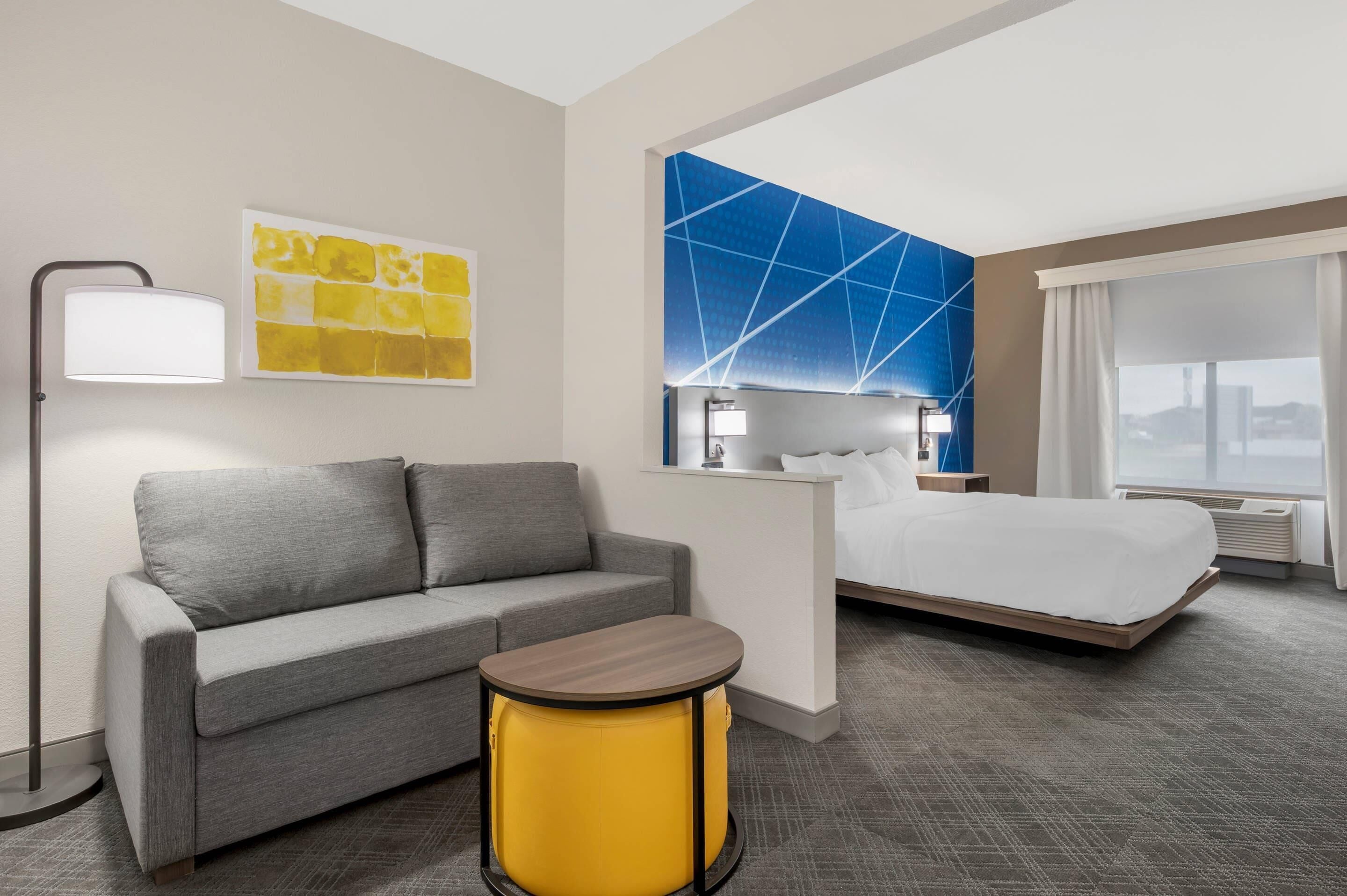Quarto de hotel com sofá cinzento, uma mesinha amarela pequena, uma cama grande, uma obra de arte geométrica azul na parede e uma mesinha de cabeceira branca, além de um abajur.