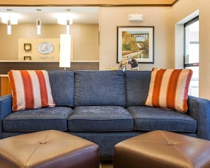 Um lobby de hotel moderno com um sofá azul, dois pufes de couro marrom, um quadro com uma imagem de uma ponte e uma placa com a inscrição 'COMFORT'.