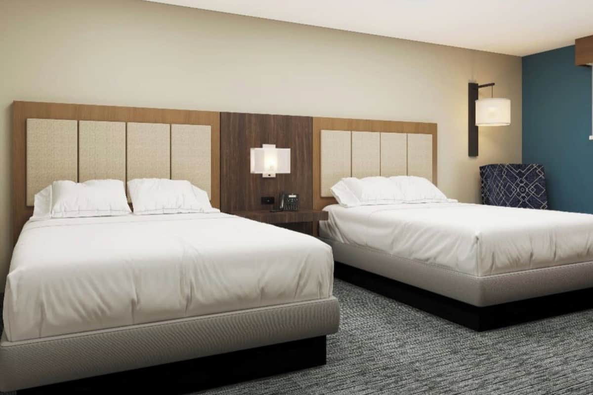 Quarto de hotel com duas camas, uma mesa de cabeceira com abajur e uma parede azul decorativa.