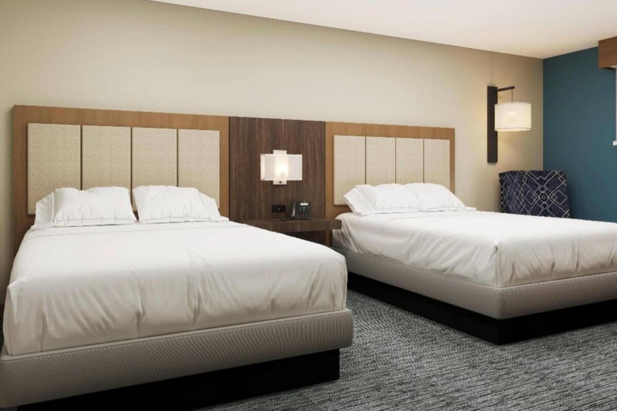Quarto de hotel com duas camas, uma mesa de cabeceira, uma luminária e um ponto de luz na parede.