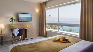 Quarto de hotel com cama, escrivaninha, cadeira, televisão, janela com vista para o mar e bandeja de frutas sobre a cama.