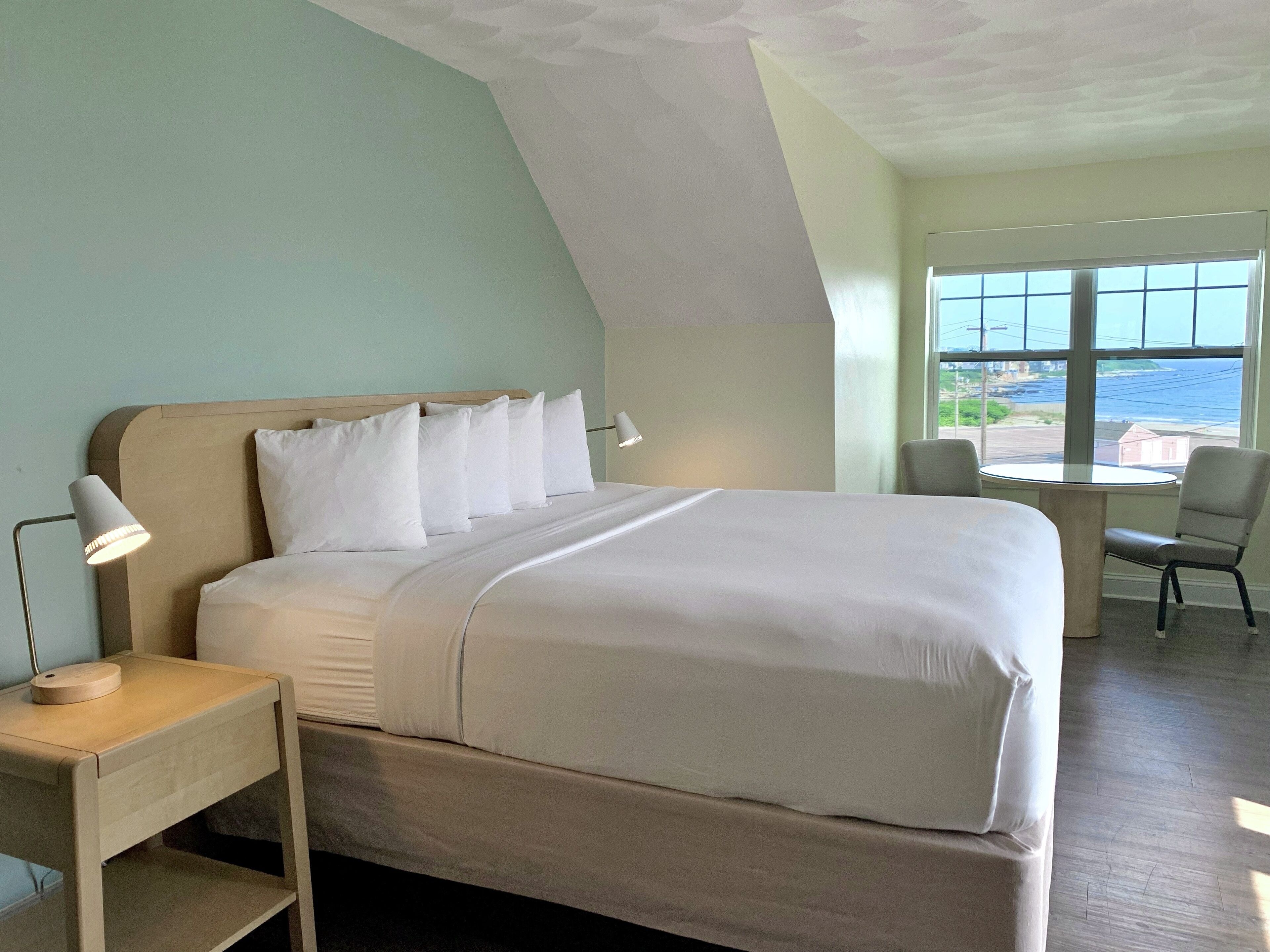 Quarto bem arrumado com cama, mesa de cabeceira, abajur e vista para o exterior através de uma grande janela.