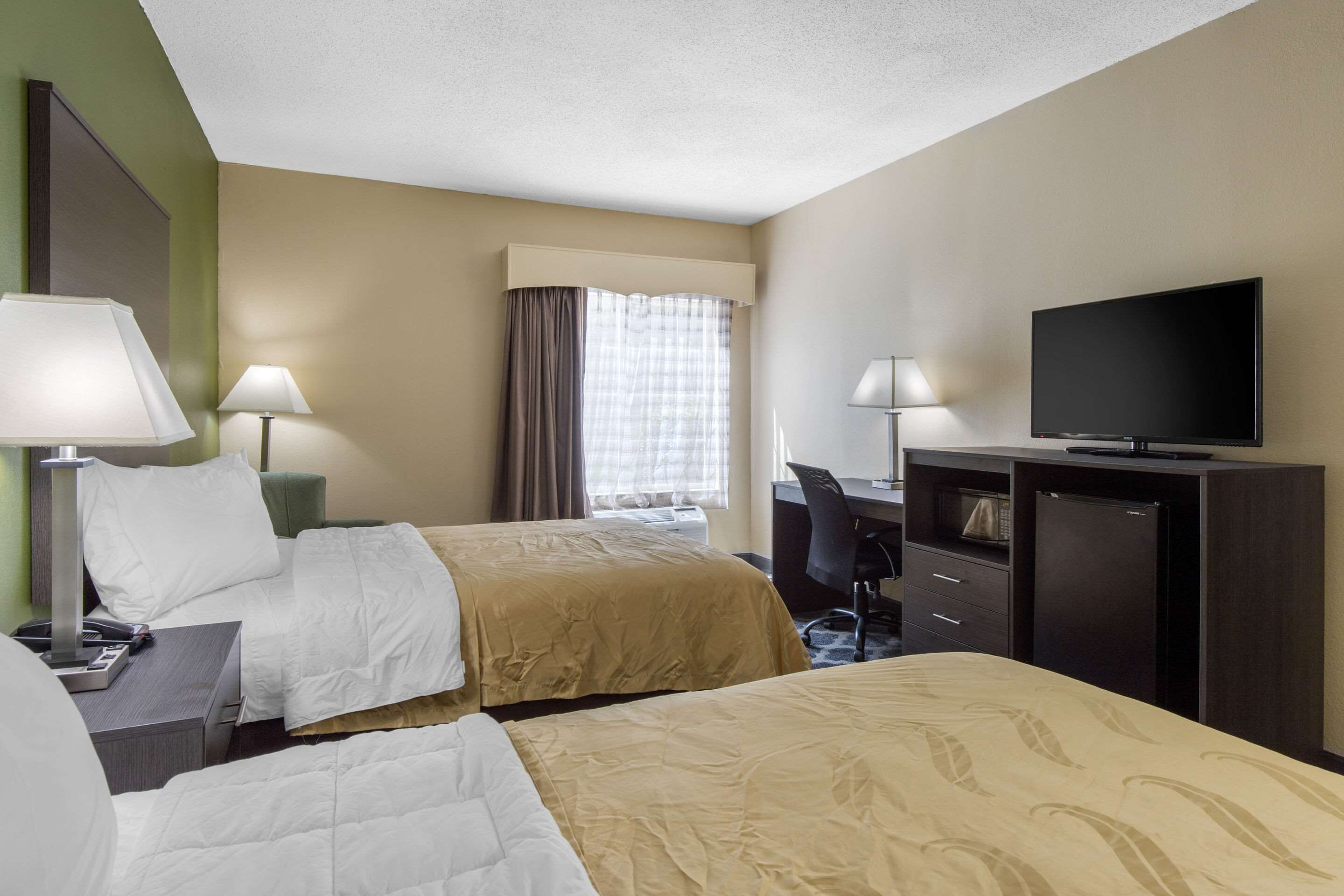 Quarto de hotel com duas camas, uma escrivaninha com computador, televisão e janela com cortinas.