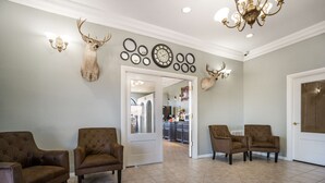 Um hall espaçoso com teto alto, um candelabro e uma parede decorada com chifres de veado e detalhes circulares. Quatro poltronas de couro marrom estão dispostas no espaço.