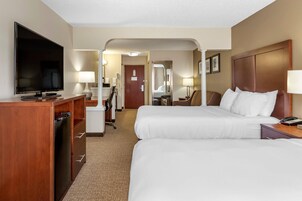 Quarto de hotel com duas camas, uma televisão, uma secretária com abajur e um banheiro visível por um arco.