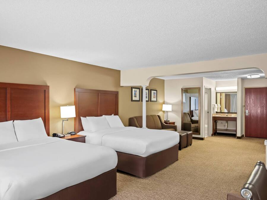 Quarto de hotel com duas camas, um espaço para sentar, uma escrivaninha, um espelho e um banheiro visível através de uma porta aberta.