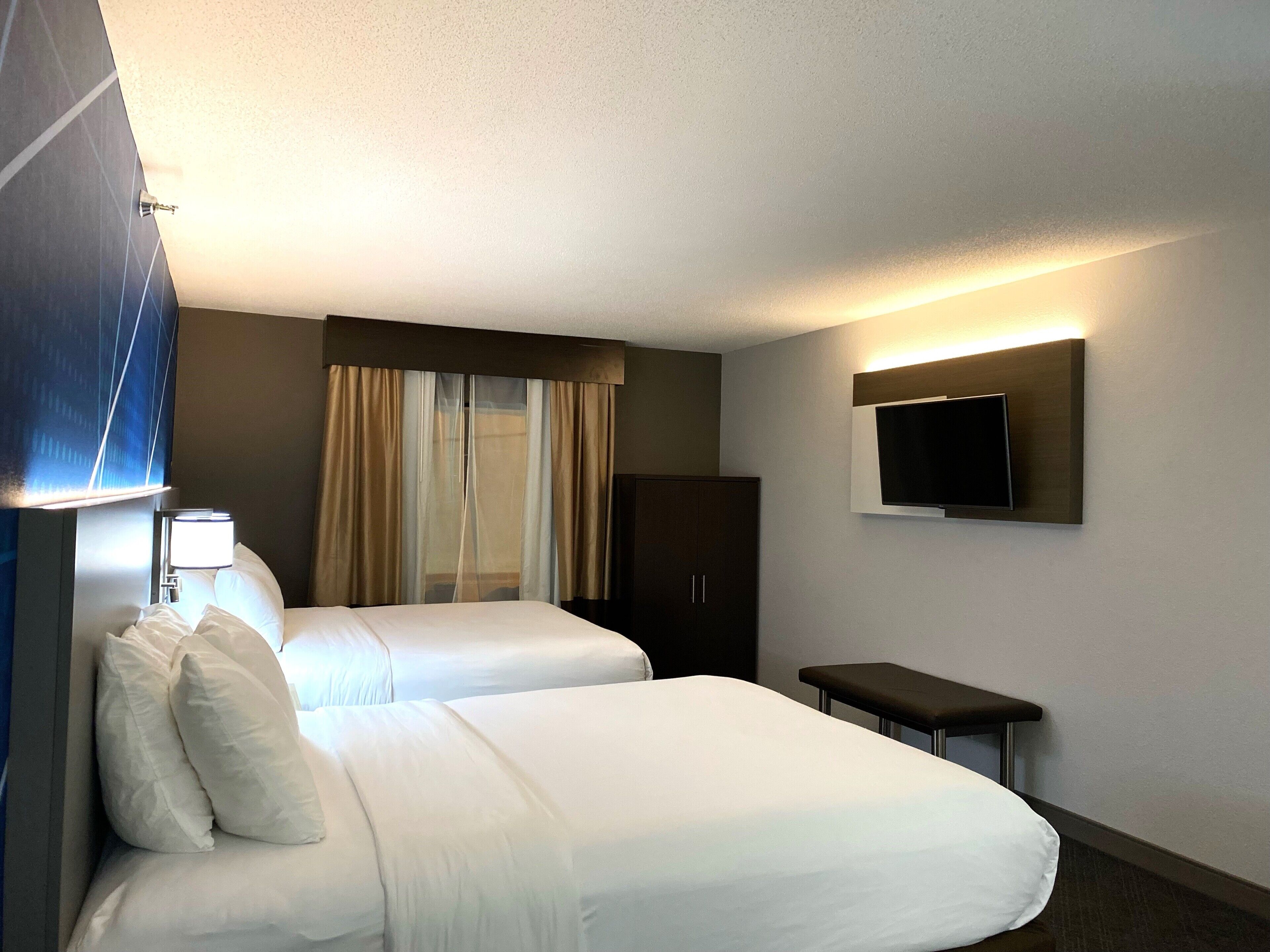 Quarto de hotel com duas camas, televisão fixada na parede, escrivaninha e janela com cortinas.