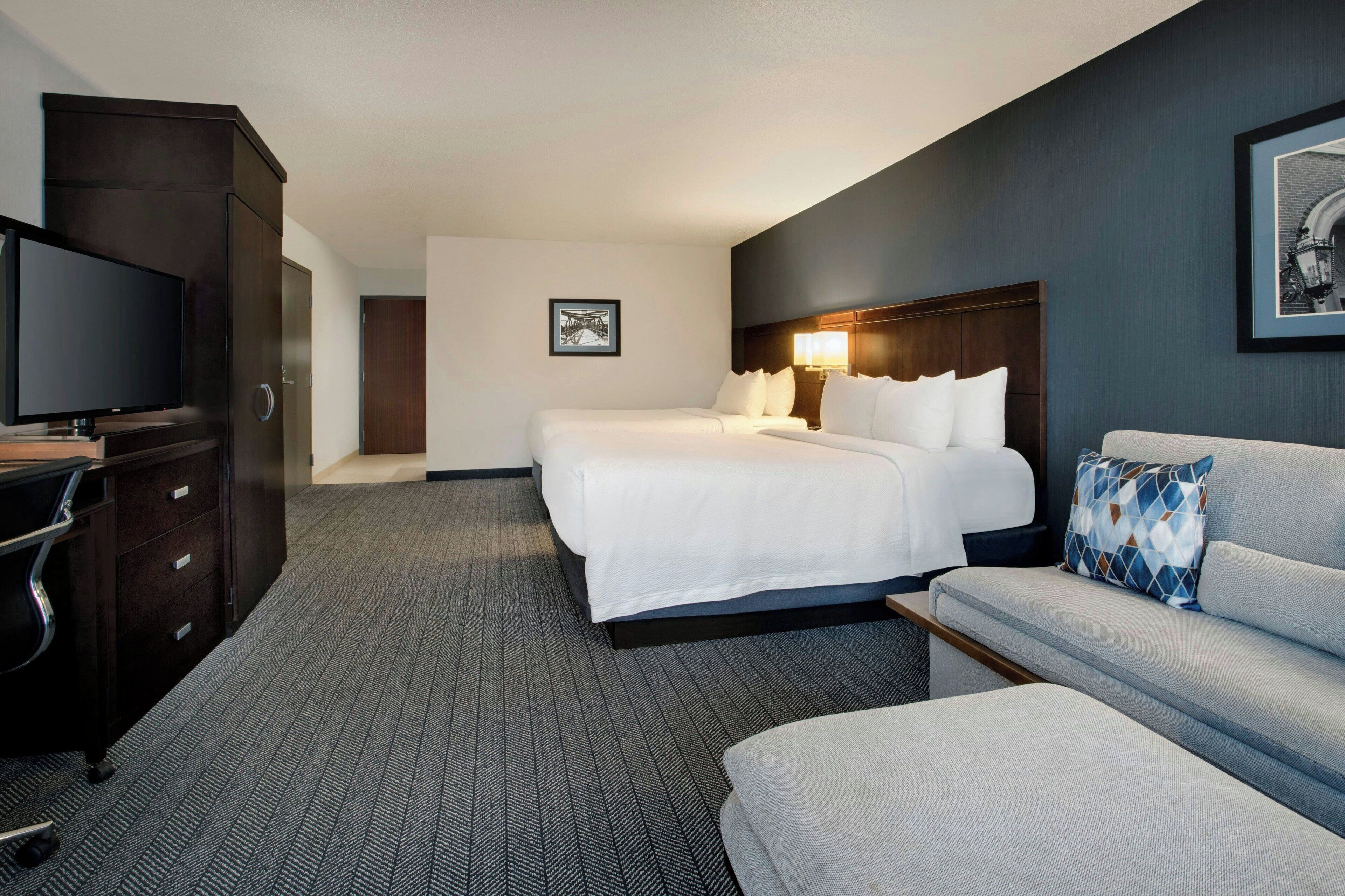 Quarto de hotel com uma cama grande, uma cama menor, um sofá, uma escrivaninha com televisão e um quadro na parede.