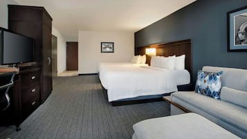Quarto de hotel com uma cama grande, uma cama menor, um sofá, uma escrivaninha com televisão e um quadro na parede.