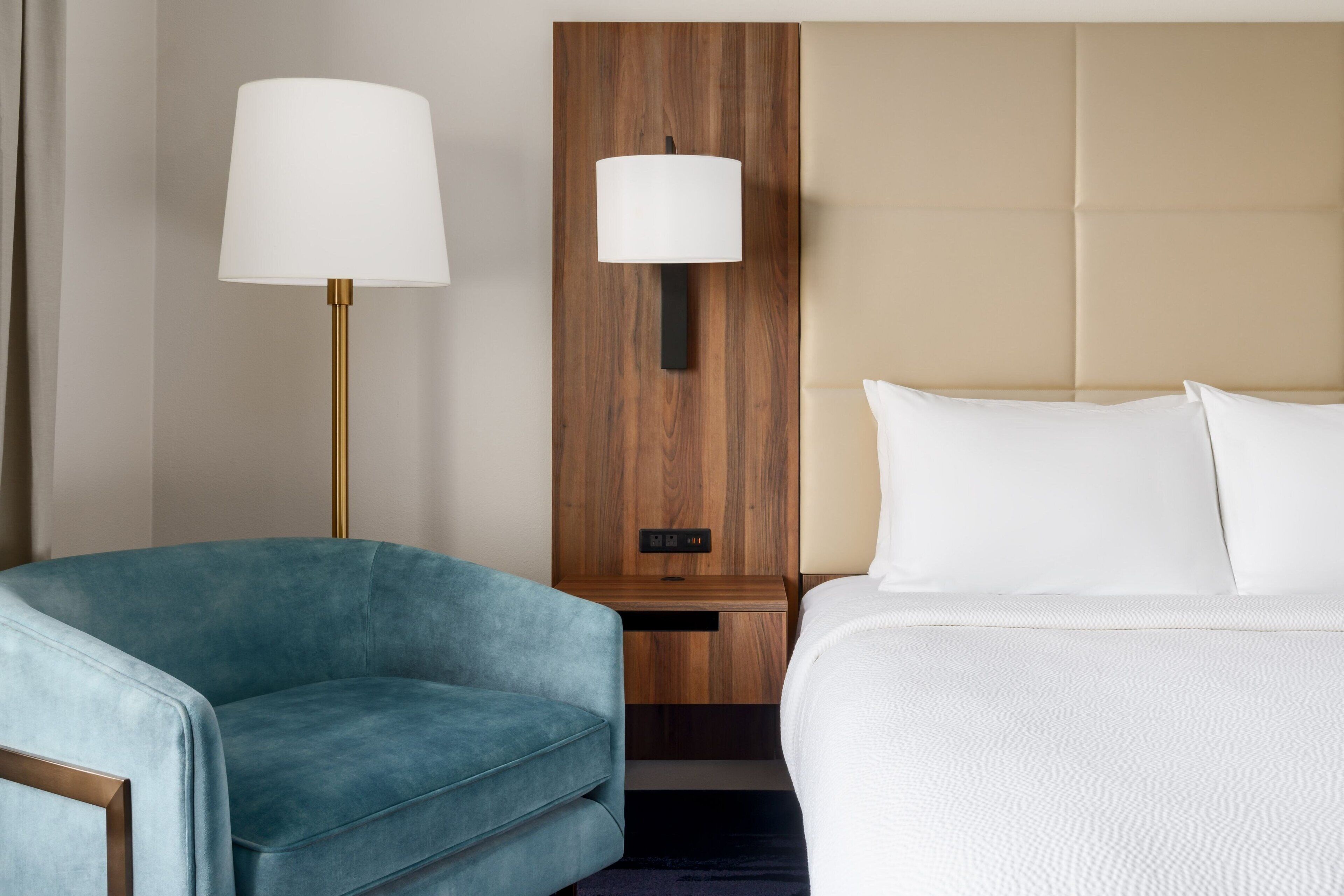 Quarto de hotel com cama, poltrona azul, painel de cabeceira de madeira e luminária fixada na parede.