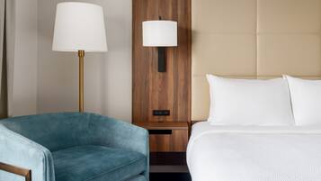 Quarto de hotel com cama, poltrona azul, painel de cabeceira de madeira e luminária fixada na parede.