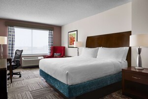 Quarto de hotel com uma cama grande, uma escrivaninha com cadeira, uma poltrona vermelha e uma janela com cortinas.