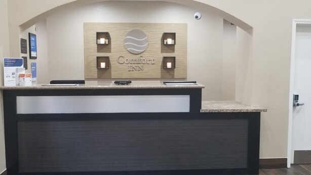 Um balcão de recepção com uma placa de madeira que diz "Comfort Inn". Há duas luminárias na parede acima da placa, uma câmera de segurança acima da porta e uma placa com a inscrição "Apenas para funcionários" na porta.