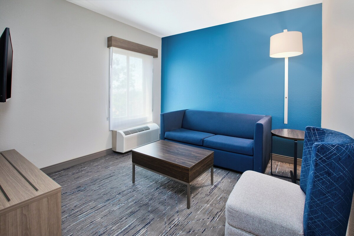 Quarto de hotel moderno com parede azul de destaque, sofá, mesa de centro, luminária e televisão.