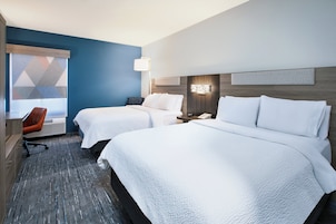 Quarto de hotel com duas camas, uma escrivaninha, uma cadeira e uma janela com cortina geométrica.