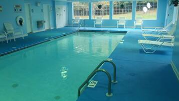 Piscina interna com água azul cristalina, cadeiras brancas na beira da piscina e vista para árvores lá fora.
