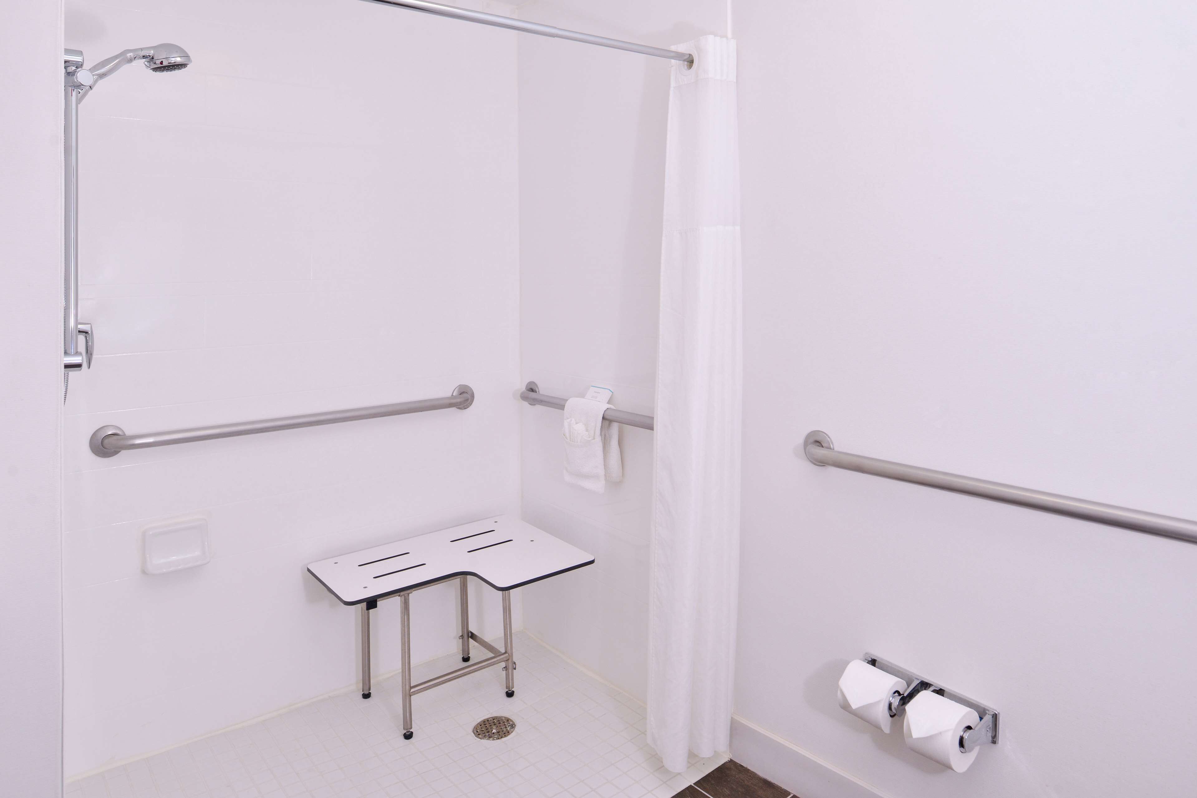 Casa de banho com chuveiro, barras de apoio, uma pequena mesa e suporte para papel higiénico.