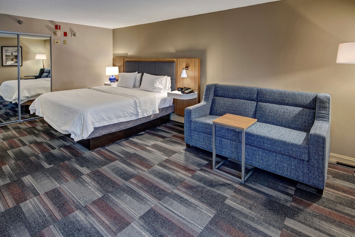 Quarto de hotel com uma cama grande, um sofá azul, uma mesinha e uma mesa de cabeceira com luminária.