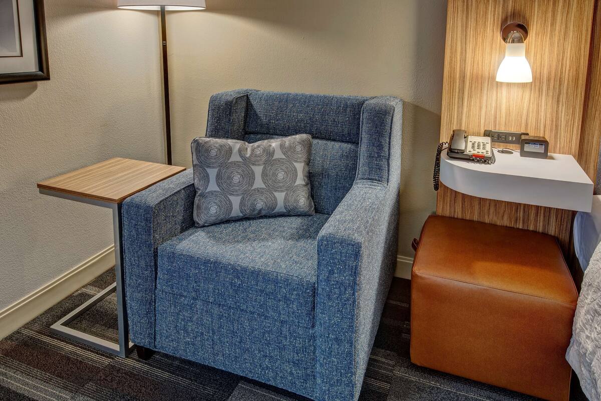 Um quarto com uma poltrona azul, uma mesinha de madeira, um banquinho marrom, uma escrivaninha com um telefone e um relógio digital, e uma luminária fixada na parede.