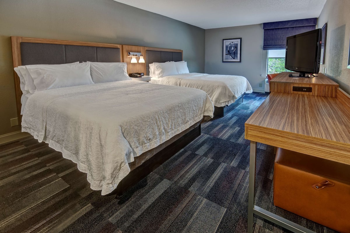 Quarto de hotel com duas camas, uma escrivaninha, uma televisão e janela com vista.