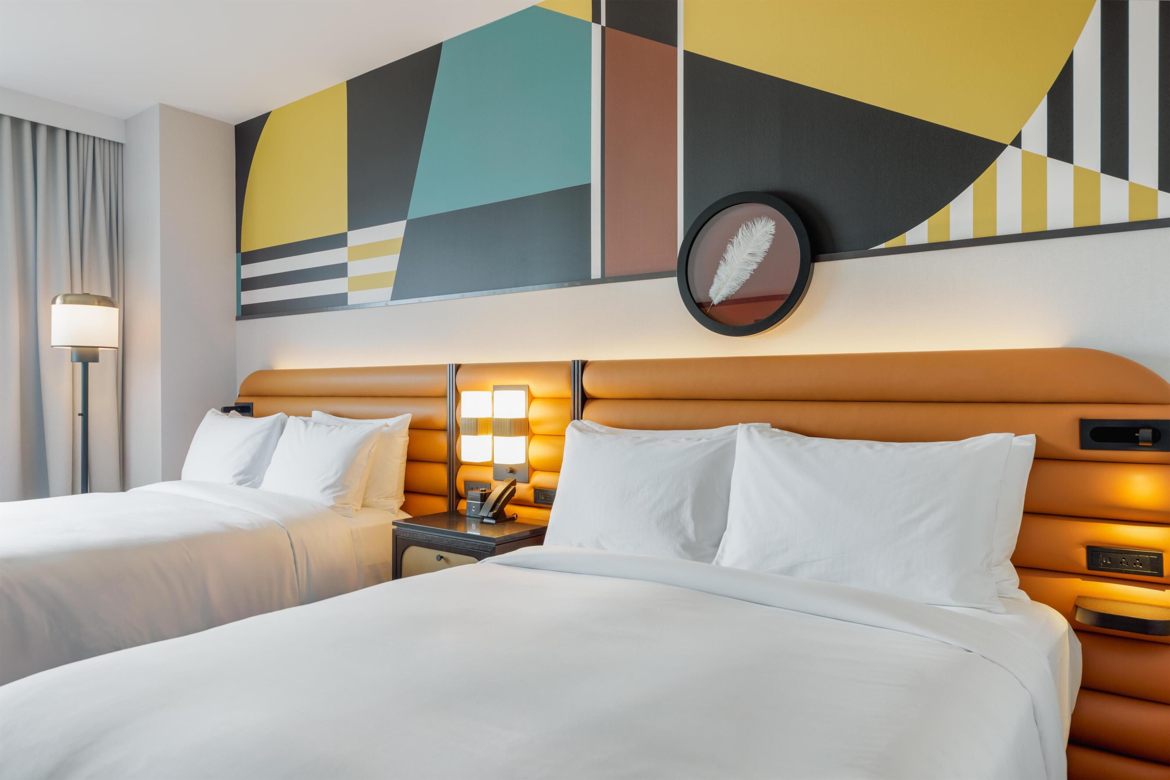 Quarto de hotel com duas camas, cabeceira com iluminação integrada, mesa de cabeceira com abajur e uma obra de arte na parede.