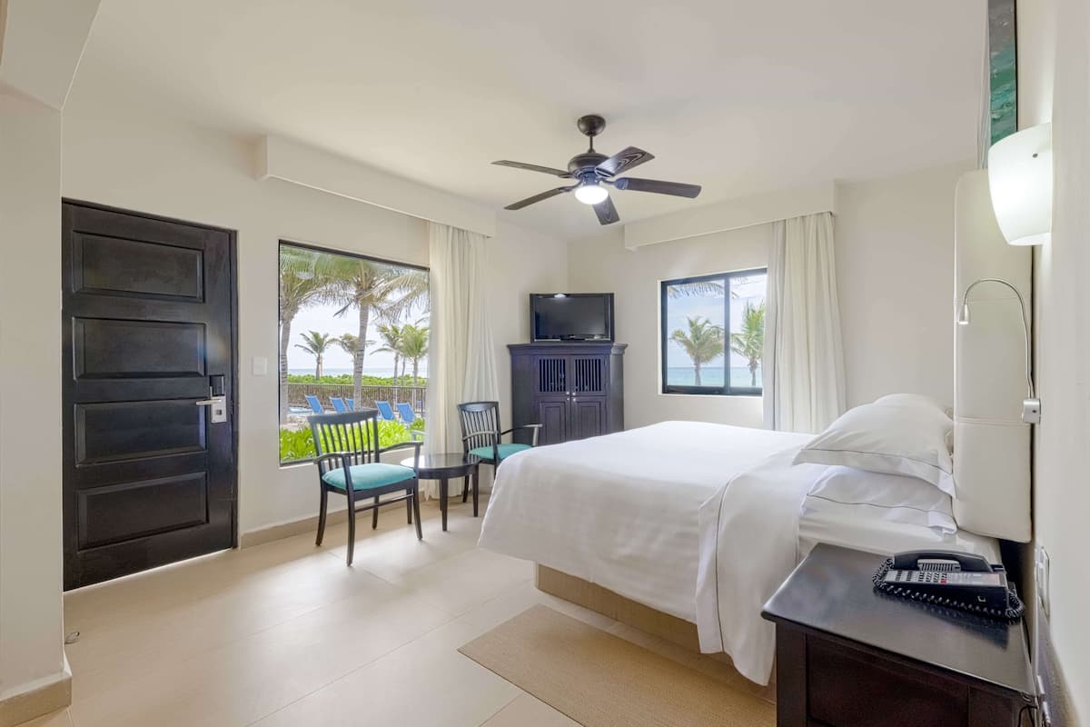 Quarto de hotel com cama, escrivaninha com telefone, televisão, ventoinha de teto e vista para o exterior.