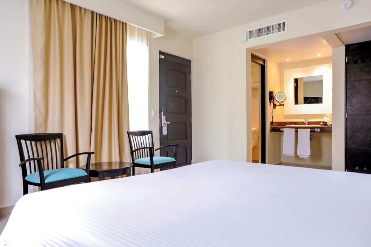 Quarto de hotel com uma cama grande, duas cadeiras, uma mesinha, banheiro com espelho e uma porta.