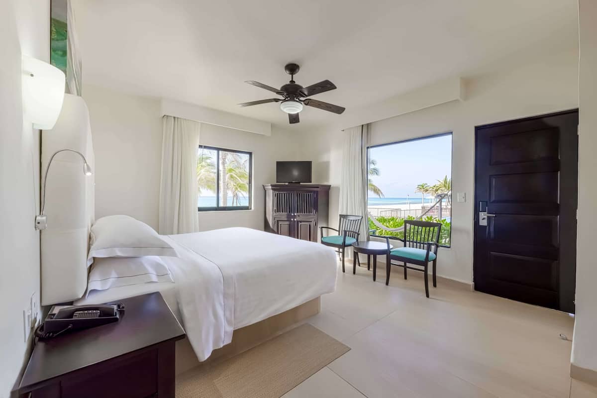 Quarto de hotel com cama, escrivaninha com luminária, televisão e varanda com cadeiras.