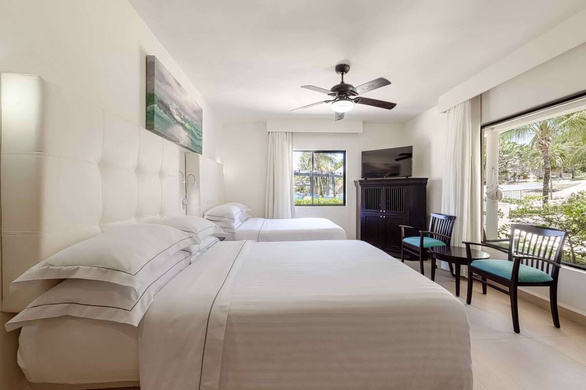 Quarto de hotel com duas camas, ventilador de teto, televisão e varanda com cadeiras.