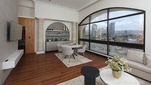 Uma sala de estar moderna com área de jantar, janelas amplas com vista para a cidade e uma TV de tela plana na parede.