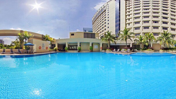 Piscina com espreguiçadeiras e palmeiras, cercada por um edifício de hotel com vários andares e design moderno.