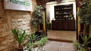 Entrada de um spa com uma placa indicando "AlammiSpa", cercada por plantas em vasos e uma parede de pedra.