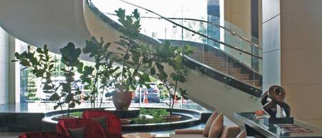 Um hall com escadaria sinuosa, assentos em vermelho e cinza, e uma planta central.