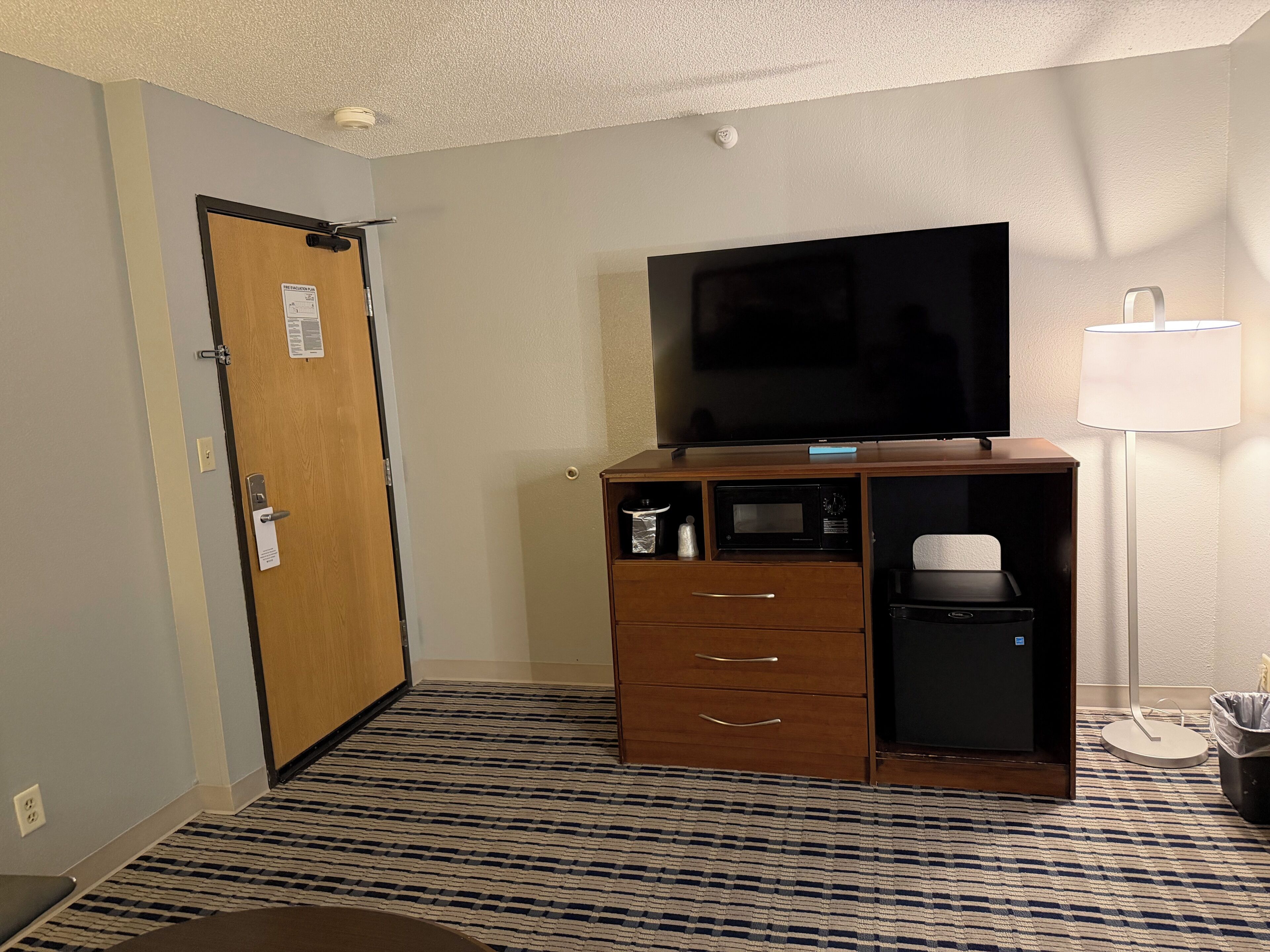 Quarto de hotel com um armário de madeira, televisão de tela plana, micro-ondas, lixeira e uma luminária.